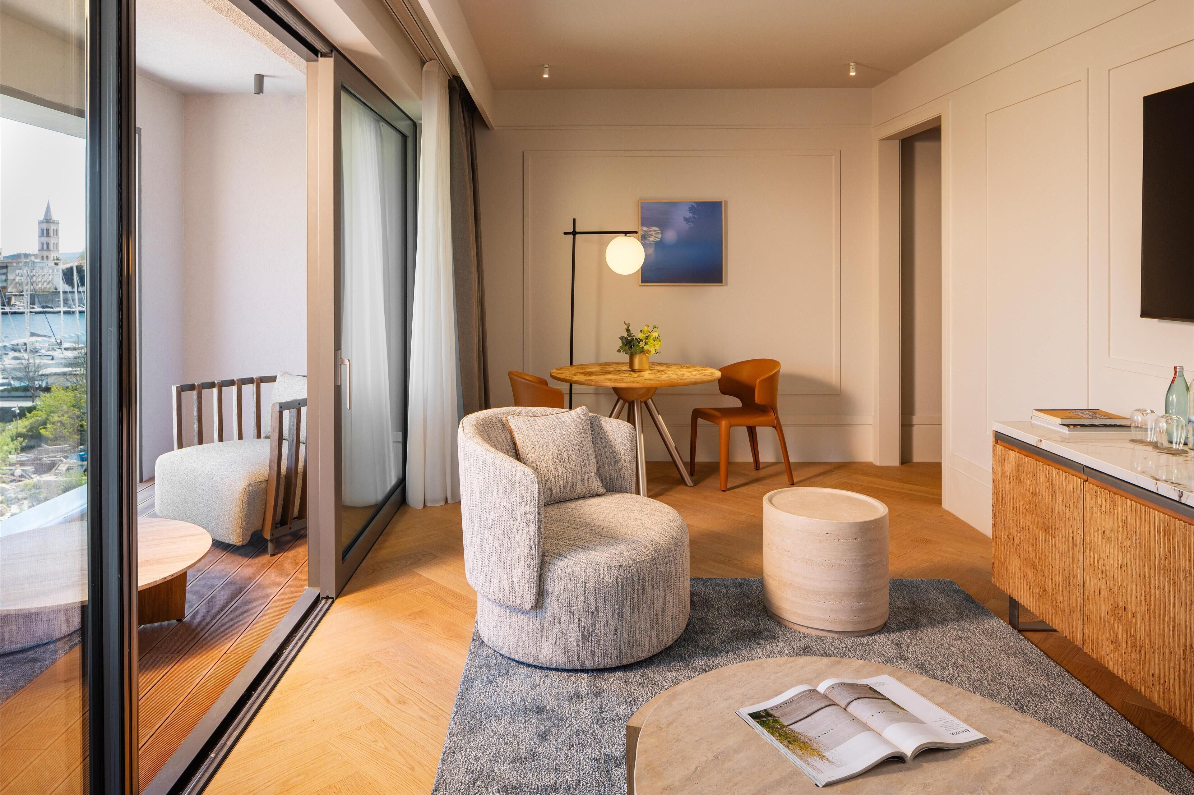 Interior moderno com área de jantar, sofá, mesa redonda e vista para a paisagem urbana através de amplas janelas.