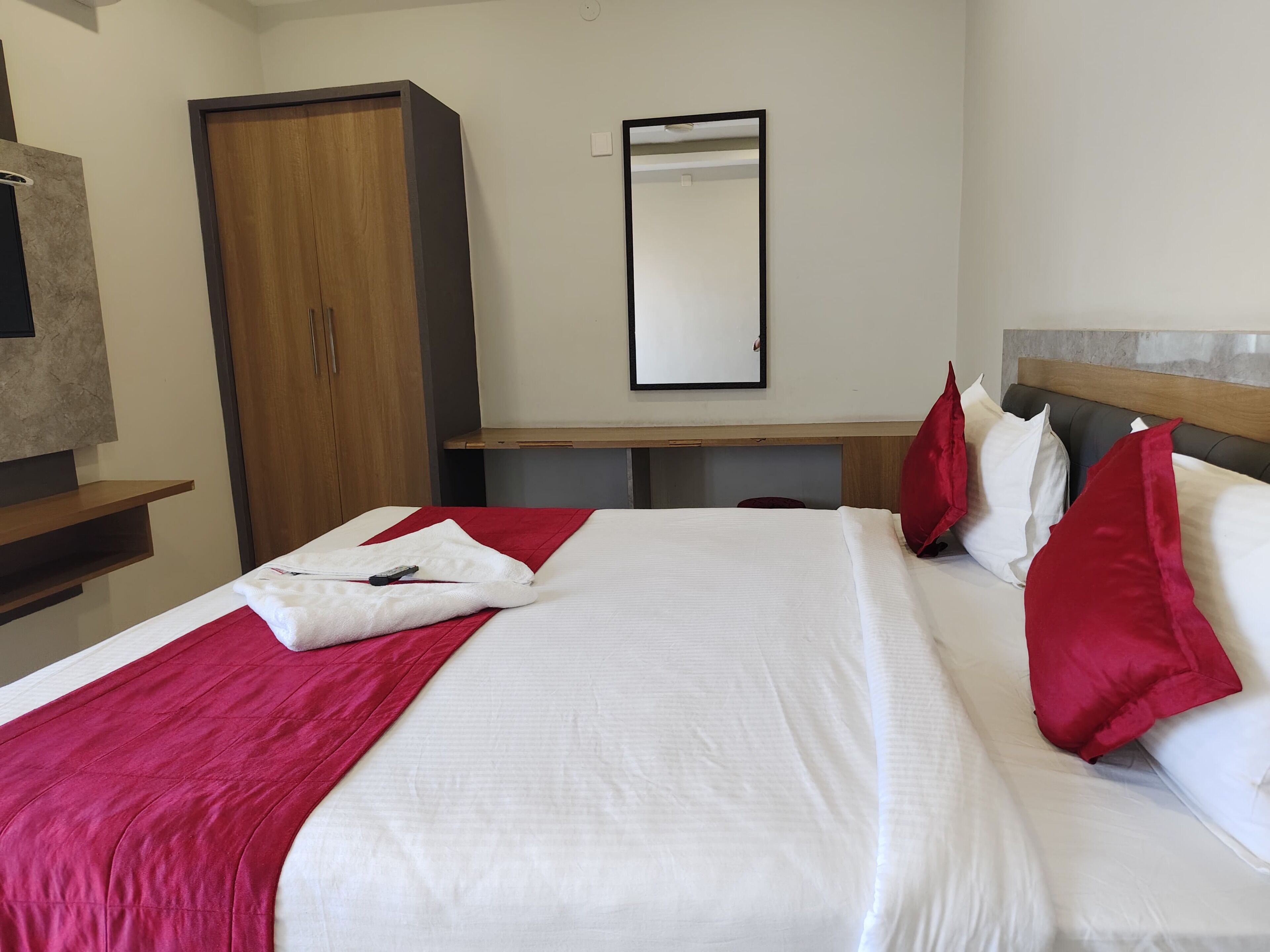 Uma cama bem arrumada com roupa de cama branca e vermelha, um guarda-roupa de madeira, uma escrivaninha pequena e um espelho na parede.