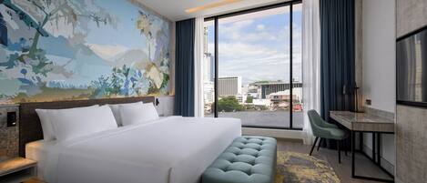 Quarto de hotel moderno com uma cama grande, uma mesa de trabalho e vista para a paisagem urbana.