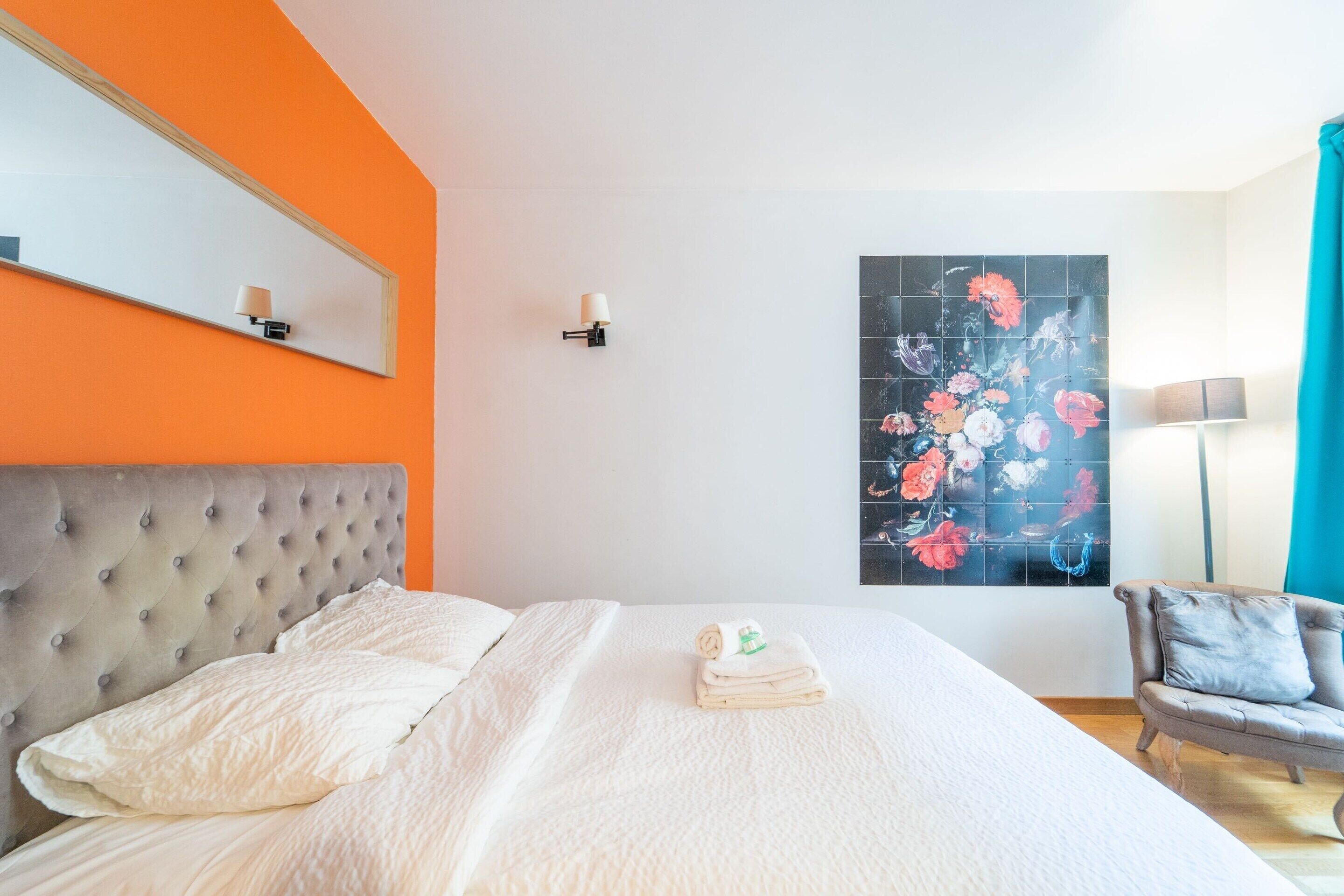 Quarto com cabeceira estofada, cama com roupa de cama branca, quadro floral na parede e mesa de cabeceira com abajur.