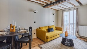 Uma sala de estar moderna com um sofá amarelo, vigas de madeira no teto, uma mesa de jantar posta com pratos e copos, e uma grande janela com cortinas.