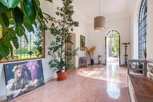 Um corredor com uma grande planta em vaso, um quadro emoldurado, uma cadeira e vista através de um arco.