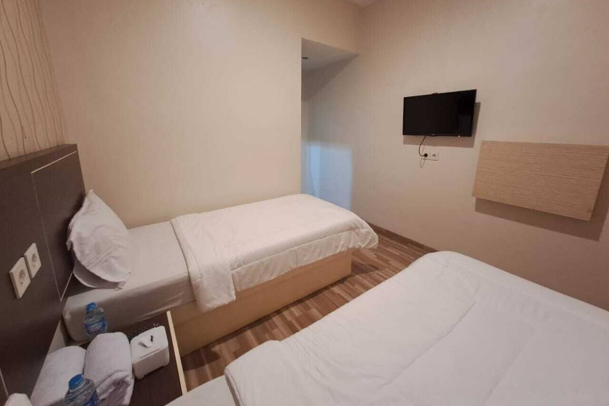 Quarto de hotel com duas camas de solteiro, televisão fixada na parede, piso de madeira e um espaço de banheiro pequeno com um porta-toalhas.
