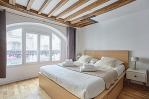 Quarto com cama de madeira, mesa de cabeceira e janela com vista para prédios.