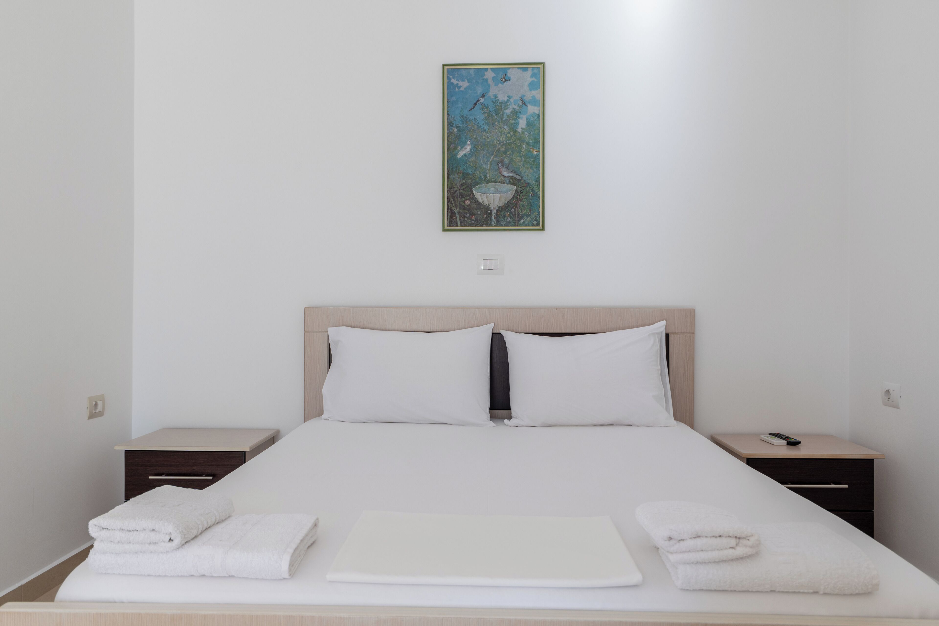 Cama bem arrumada com lençóis brancos e duas almofadas, mesa de cabeceira com controle remoto e pintura de pássaros na parede.
