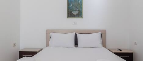 Cama bem arrumada com lençóis brancos e duas almofadas, mesa de cabeceira com controle remoto e pintura de pássaros na parede.