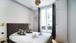 Quarto com cabeceira estofada, uma janela ampla com vista para edifícios, guarda-roupa e mesa de cabeceira com abajur.