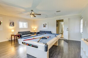 Uma mesa de bilhar com bolas, um ventilador de teto e um sofá em um ambiente com piso de madeira e paredes brancas.