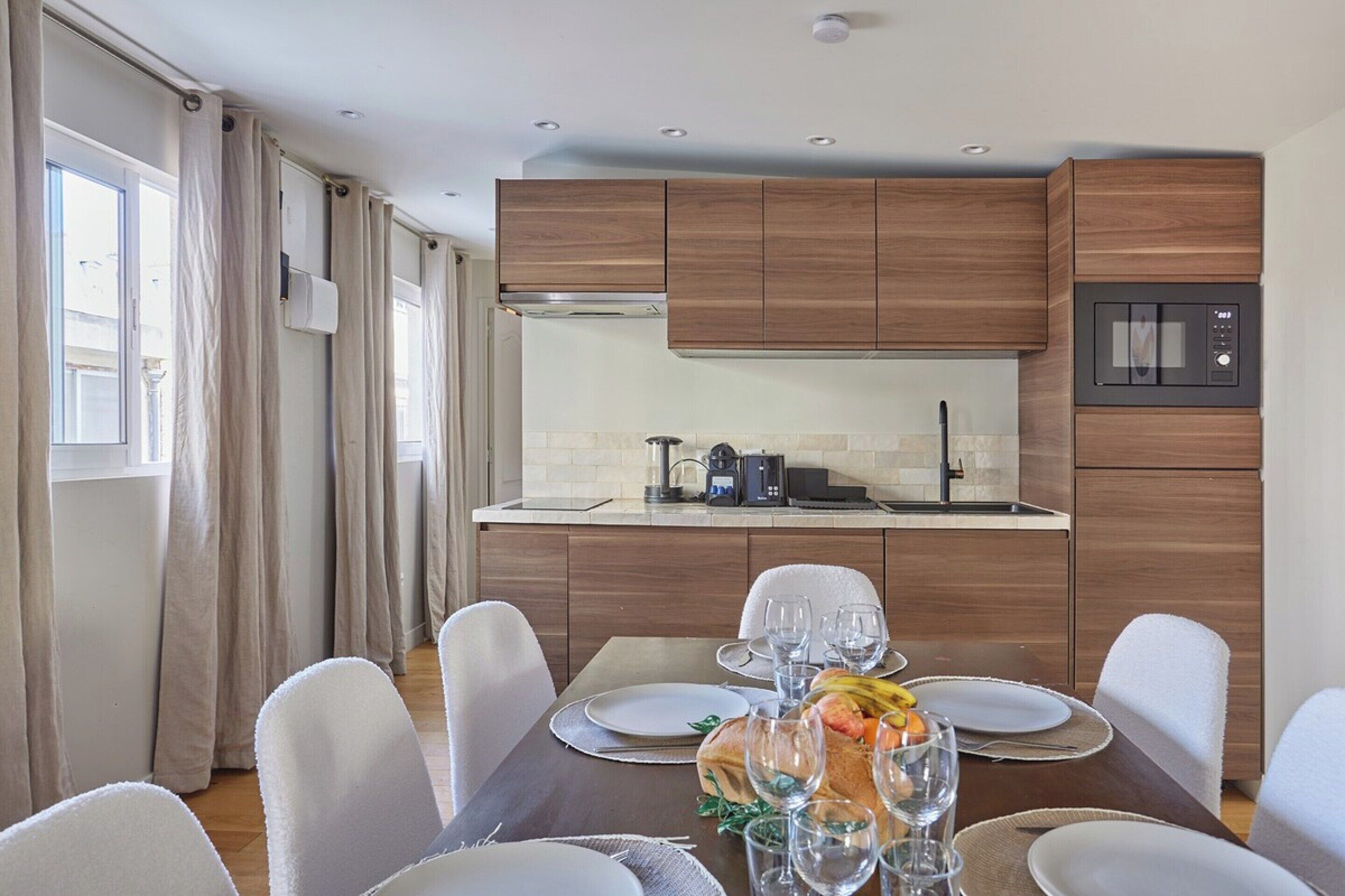 Cozinha moderna com armários de madeira, micro-ondas embutido e área de refeições para seis pessoas, com cadeiras brancas e mesa posta para uma refeição.