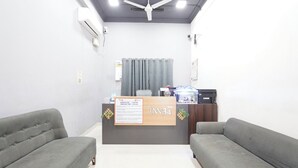 Uma área de recepção com balcão de check-in, dois sofás cinzentos, um ventilador de teto e uma pequena casa modelo no balcão.