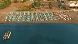 Vista aérea de uma praia com fileiras de espreguiçadeiras e guarda-sóis, um barco atracado na água e um edifício ao fundo.