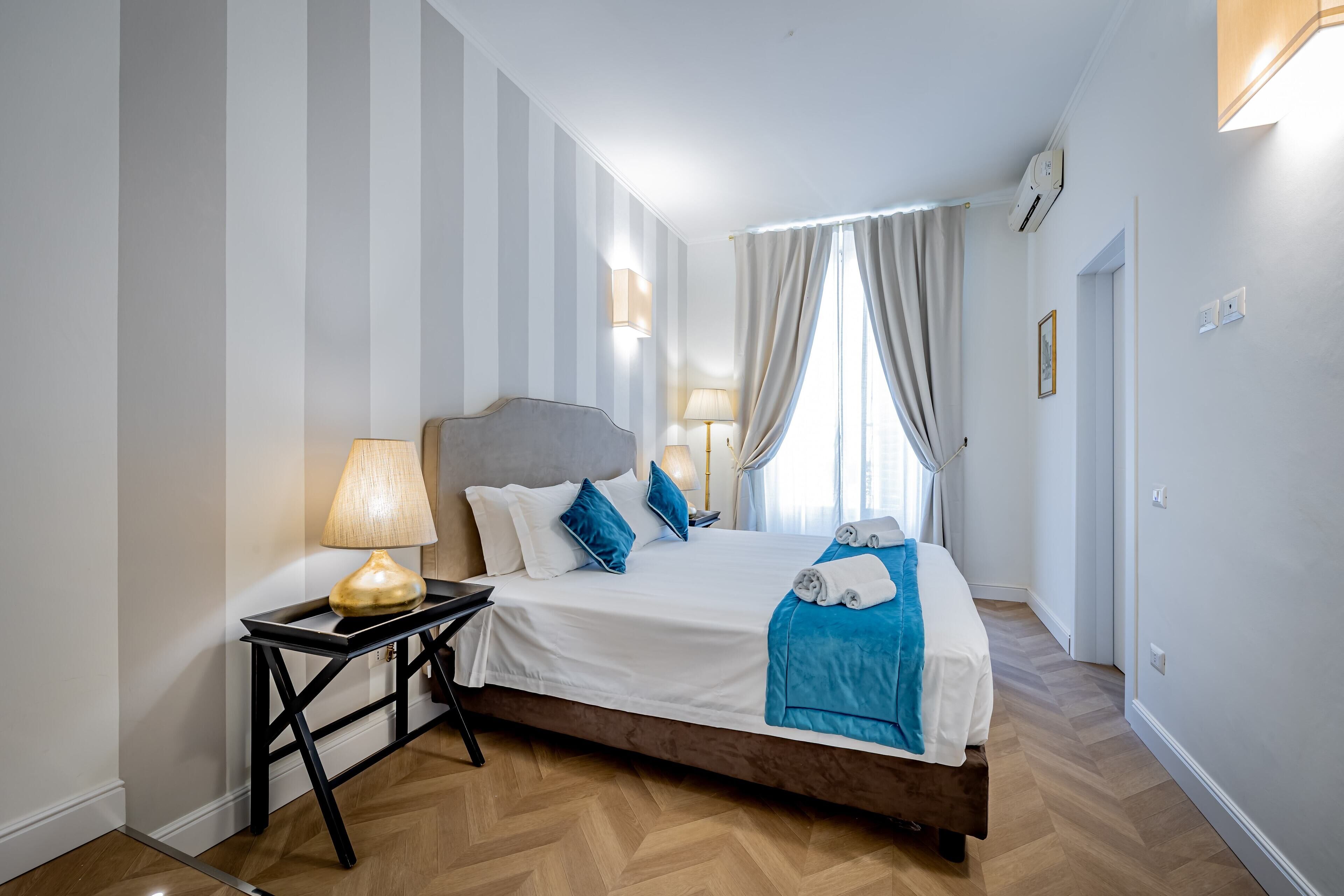 Um quarto bem arrumado com cama, mesa de cabeceira, abajur e cortinas.