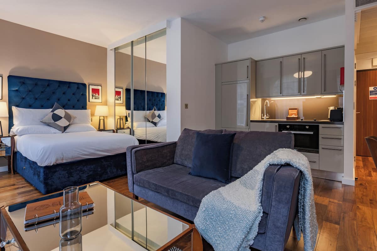 Quarto de hotel moderno com cama, sofá, mesa de centro de vidro, cozinha compacta e espelho.