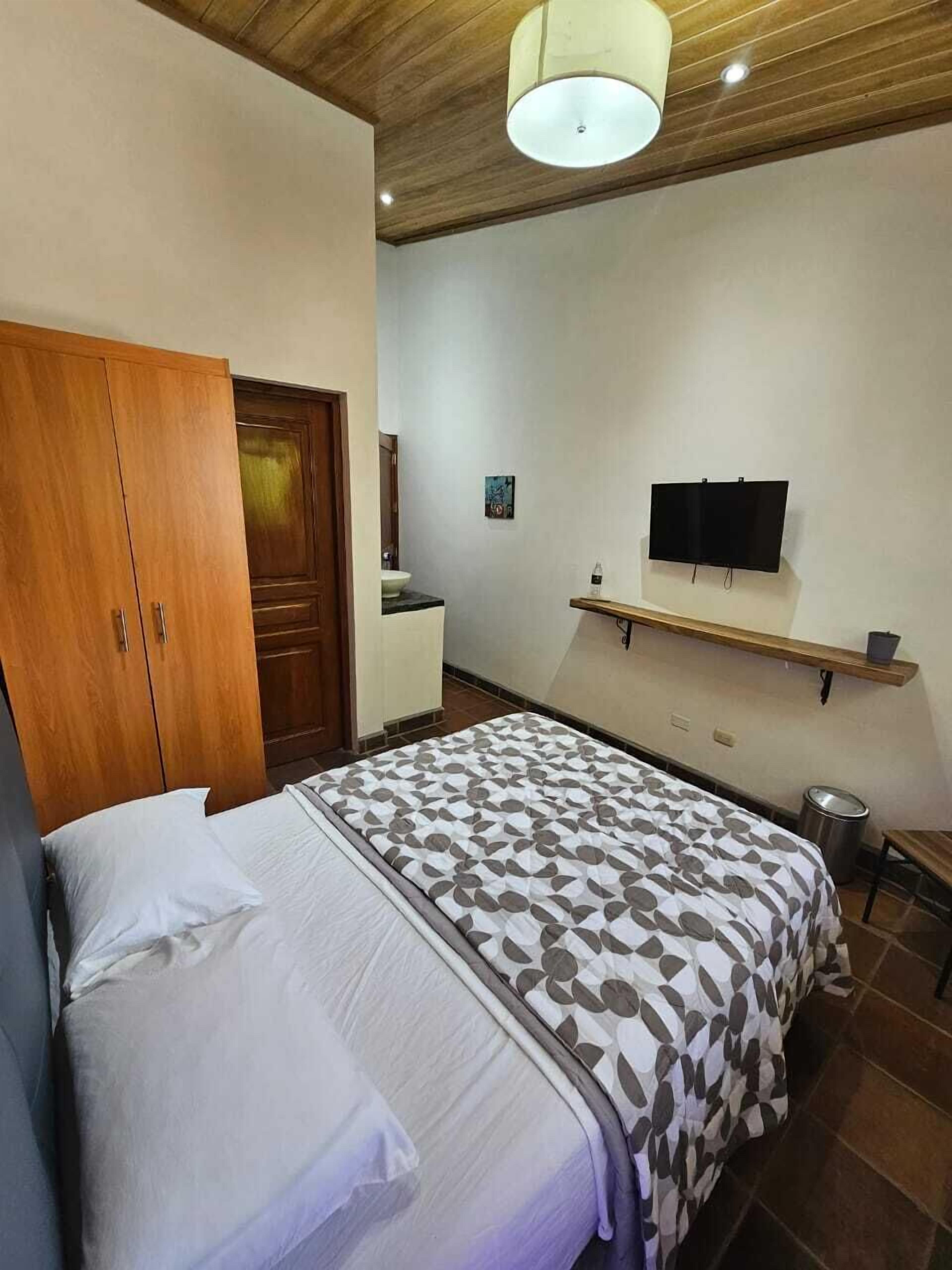 Quarto com cama, guarda-roupa de madeira, televisão na parede e uma pequena prateleira.