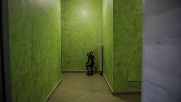 Um corredor estreito com paredes verdes e piso de madeira. Um radiador está fixado na parede, e uma pessoa senta-se numa cadeira ao centro do corredor.