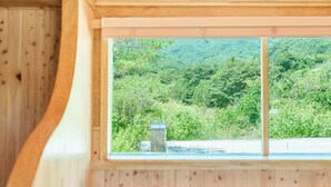 Interior de uma cabana de madeira com uma janela grande que dá para uma encosta arborizada.