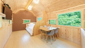 Uma cabana de madeira acolhedora, com uma cama, mesa de jantar e grandes janelas com vista para o verde.