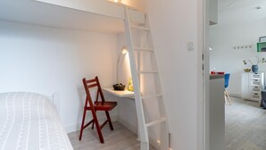 Um quarto com uma cama, uma cadeira de madeira, uma escrivaninha com abajur e um guarda-roupa branco.