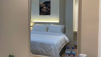 Quarto de hotel com cama, mesa de cabeceira, cafeteira, garrafas de água e espelho refletindo o ambiente.