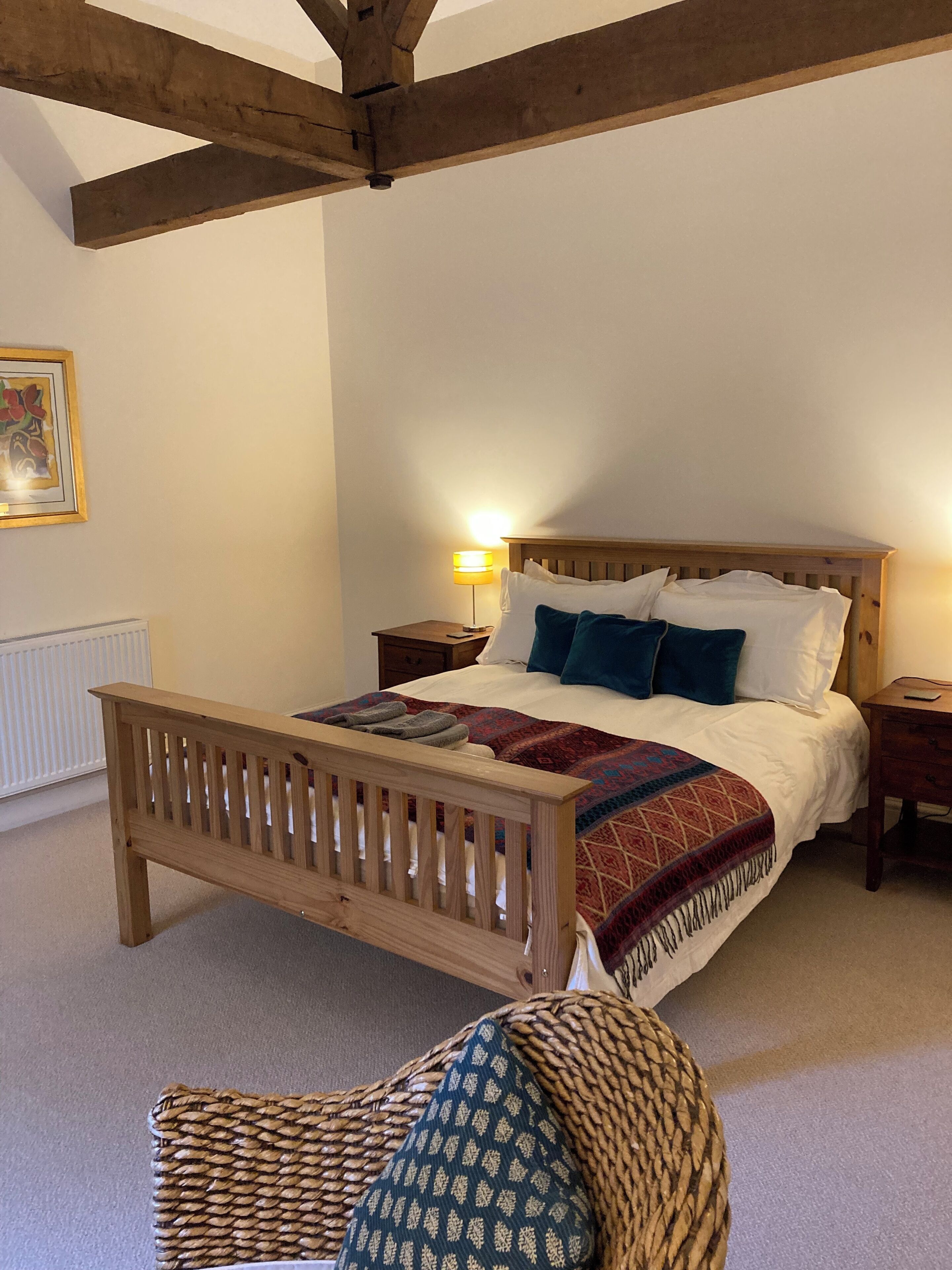 Quarto com cama de madeira, mesa de cabeceira com abajur, radiador e cadeira de fibra sintética.
