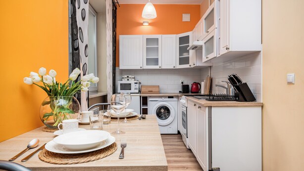 Cozinha compacta com armários brancos, máquina de lavar e área de jantar com mesa posta para dois.