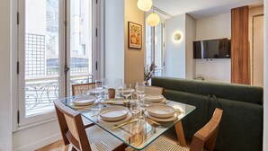 Uma sala de jantar com uma mesa de vidro posta para quatro pessoas, um sofá verde e uma TV fixada na parede.