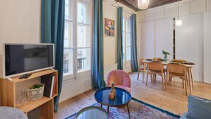 Uma sala de estar moderna com televisão, mobiliário de madeira, uma mesa de jantar e janelas amplas com cortinas.
