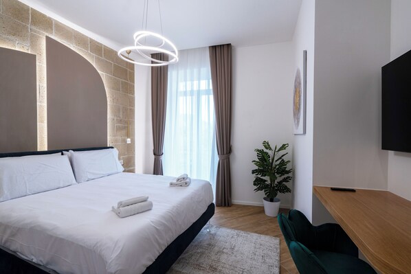 Quarto de hotel moderno com uma cama grande, uma mesa de escritório, uma planta em vaso e uma TV na parede.