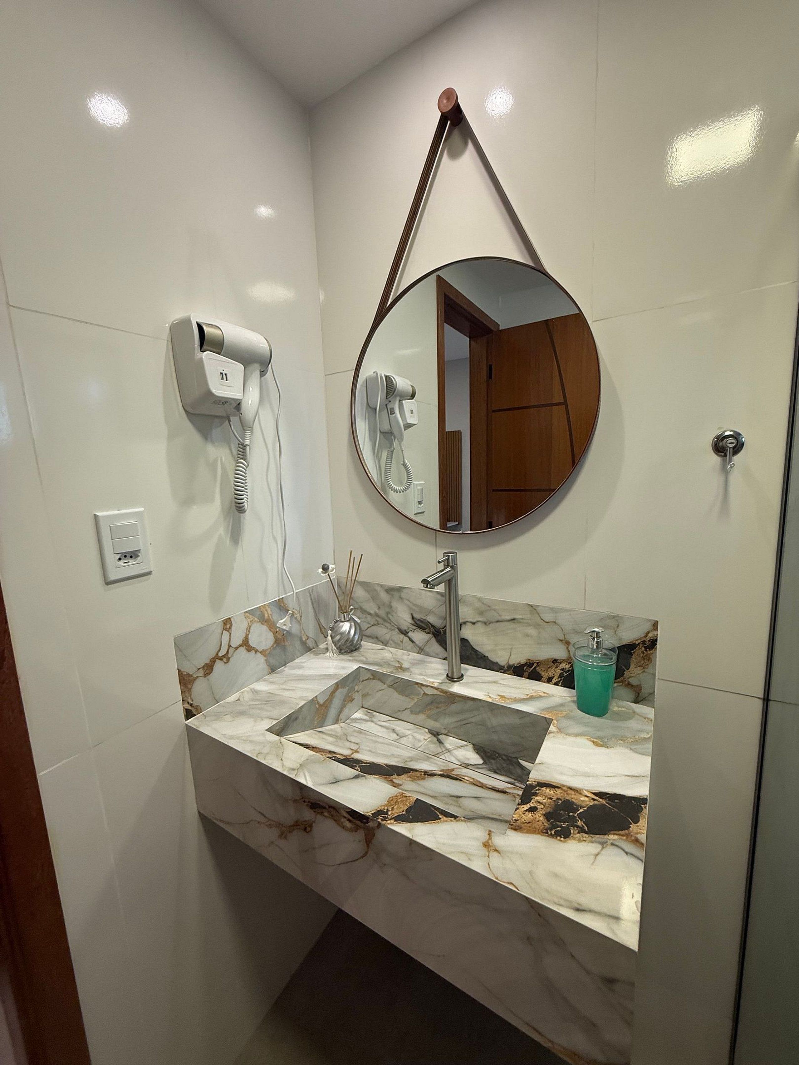 Casa de banho com lavatório de mármore, espelho redondo, secador de cabelo e dispensador de sabonete.