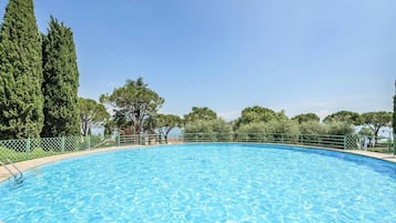 Piscina azul límpida rodeada por uma cerca verde, com árvores e um céu claro ao fundo.
