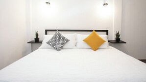 Cama bem arrumada com roupa de cama branca e duas almofadas decorativas, com duas mesinhas de cabeceira pequenas com plantas em vasos.