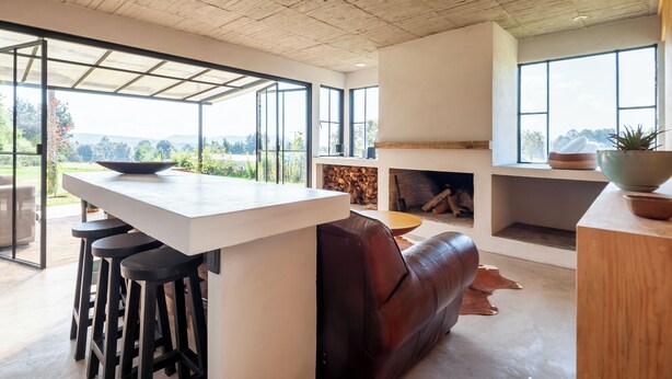 Uma cozinha moderna com uma ilha grande, banquetas de bar, lareira e janelas amplas com vista para o exterior.