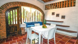 Um quarto com parede de tijolos brancos, porta de madeira, sofá azul, mesa branca com abacaxi e frutas, e chão de azulejos.