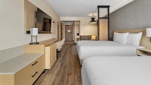 Quarto de hotel com duas camas, uma escrivaninha, uma cadeira, um aparador e um espelho grande.
