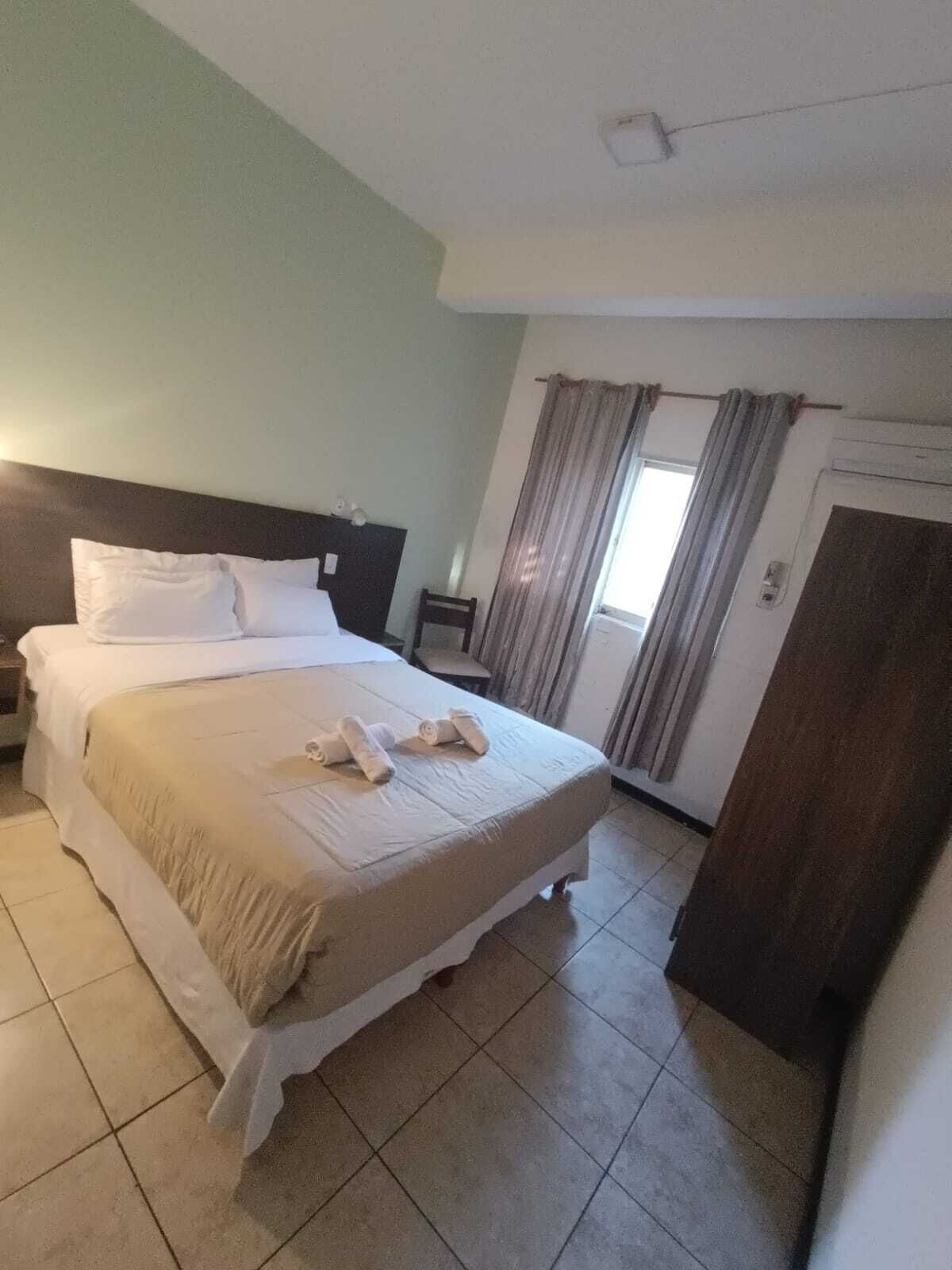 Quarto de hotel com cama, mesa de cabeceira, cadeira, janela com cortinas e porta.