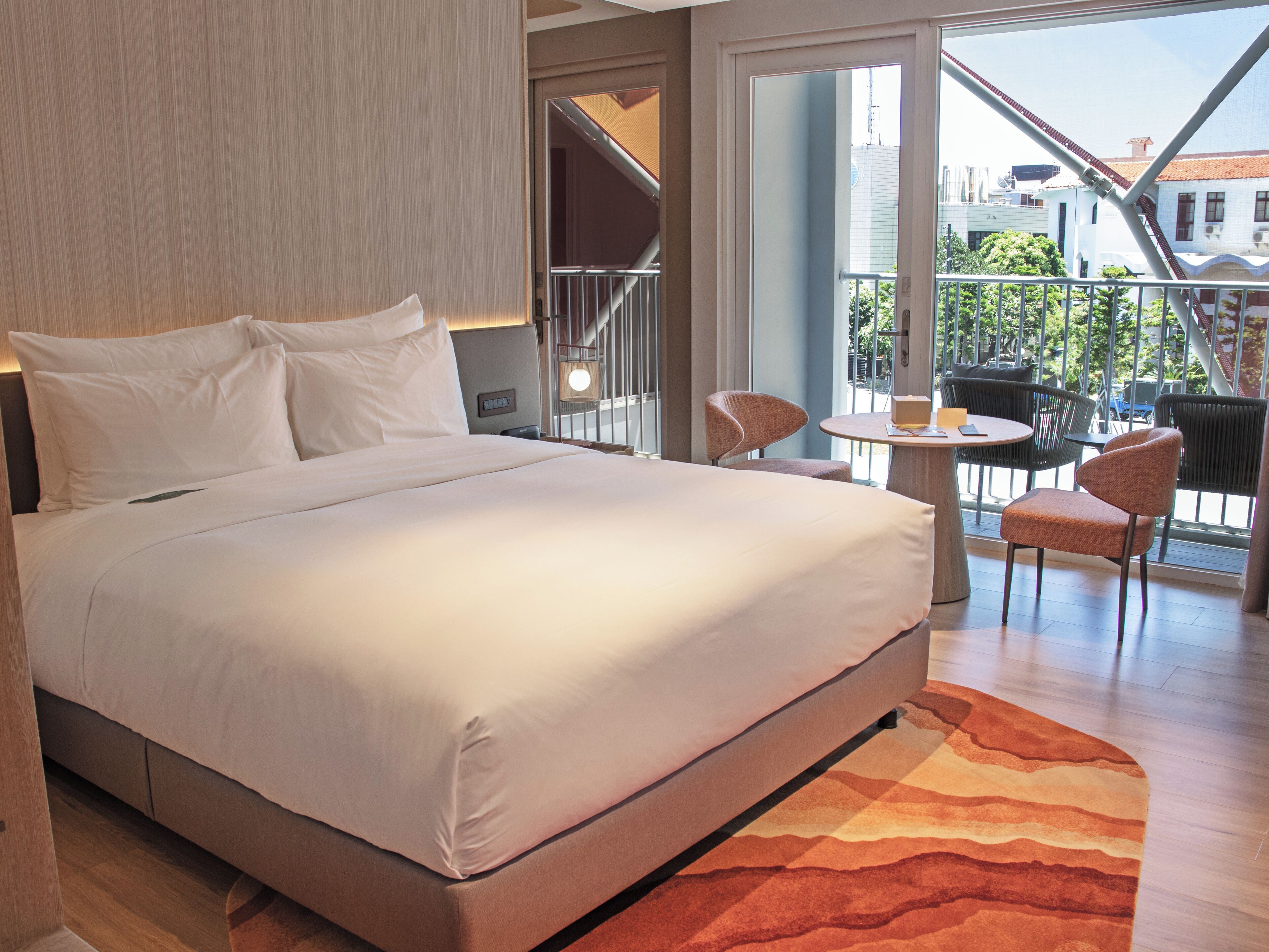 Quarto de hotel com uma cama grande, uma mesa pequena e cadeiras. Possui uma varanda com vista para edifícios e árvores.