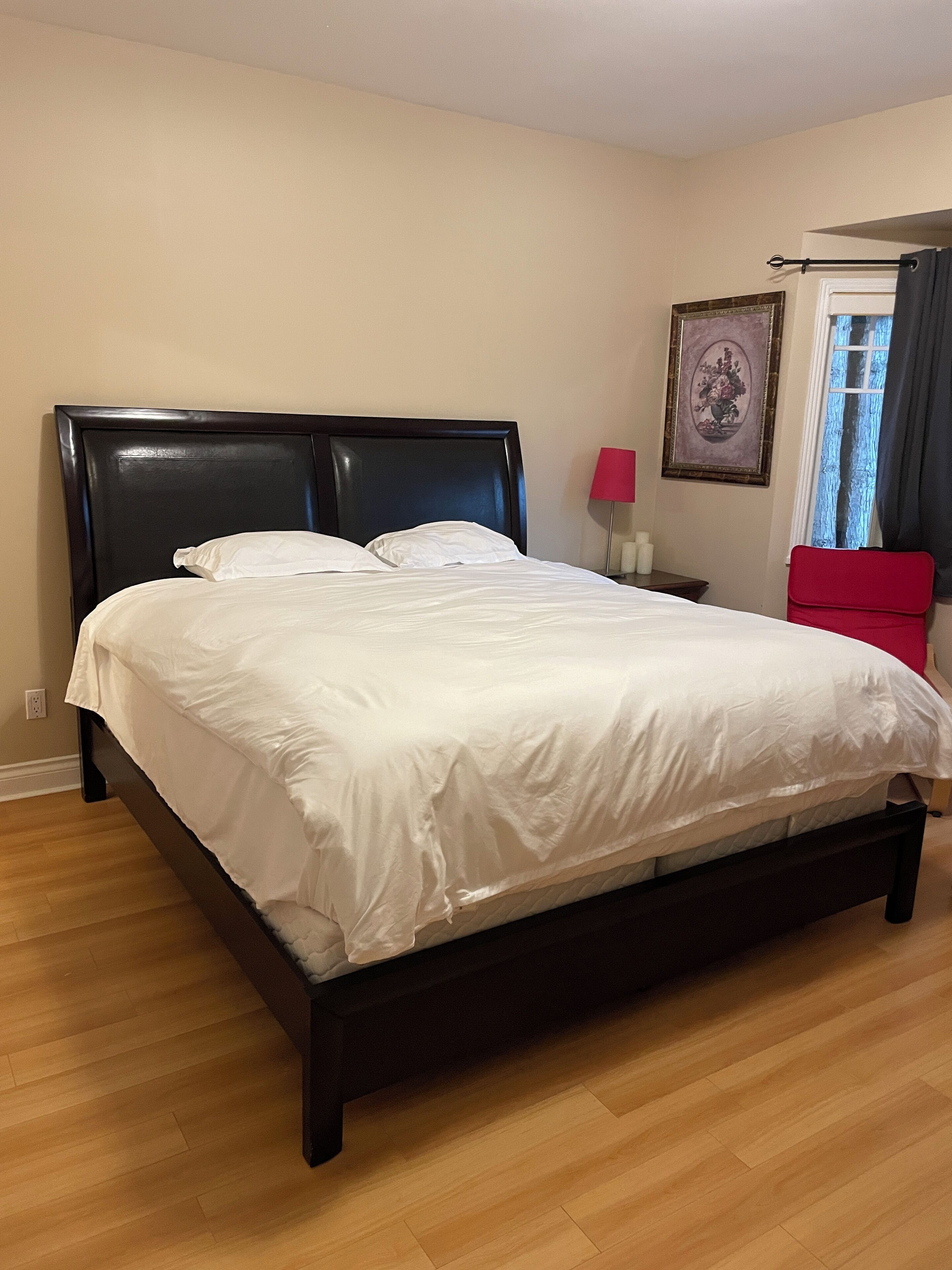 Cama bem arrumada num quarto com piso de madeira, cabeceira escura, mesinha de cabeceira com abajur e um quadro na parede.