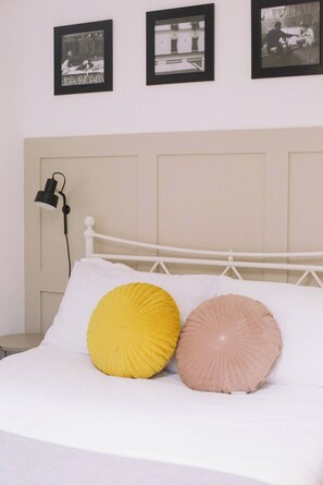 Uma cama com roupa de cama branca e duas almofadas decorativas, cabeceira branca e luminária na parede.
