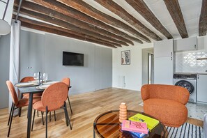 Uma área de refeições com mesa posta para dois, um sofá e uma cozinha com máquina de lavar.