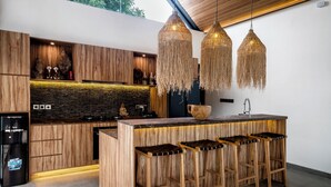 Uma cozinha moderna com armários de madeira, uma ilha central com bancos, luminárias pendentes de vime e uma parede envidraçada preta.