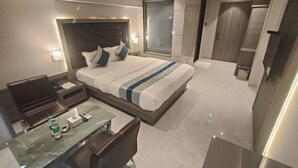 Quarto de hotel moderno com uma cama grande, uma escrivaninha com cadeira, mesa de cabeceira e banheiro com chuveiro de vidro.