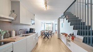 Cozinha moderna com armários brancos, bancada de madeira e cuba de aço inoxidável. Há uma área de refeições com mesa e cadeiras, e uma escada com corrimão de metal.