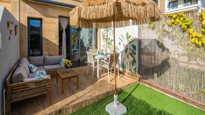Um pátio moderno ao ar livre com guarda-sol de palha, um sofá, uma mesa de jantar e uma pequena área de jardim com grama artificial e plantas.