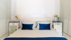 Cama bem arrumada com cabeceira branca, duas mesas de cabeceira com abajures e um pequeno armário.