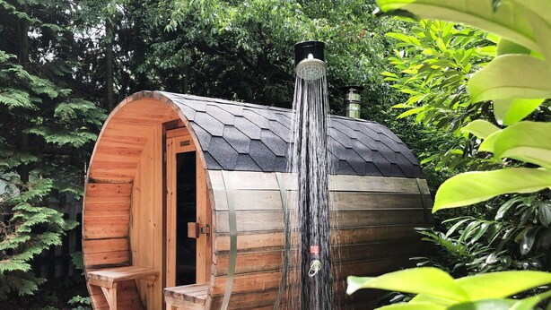 Uma estrutura em forma de barril de madeira, com uma porta e degraus que levam a entrada, rodeada por vegetação.