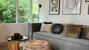 Uma sala de estar moderna com um sofá cinzento, um banquinho de vime redondo, uma mesinha redonda com uma vela e um abajur.