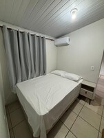 Quarto individual com uma cama de solteiro, cortinas cinzas, ar condicionado, mesa de cabeceira e piso cerâmico.