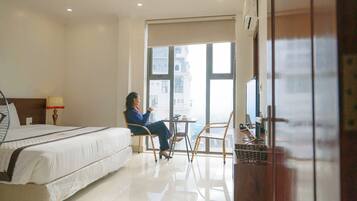 Quarto de hotel com cama, escrivaninha e cadeira. Janela com vista para edifícios.