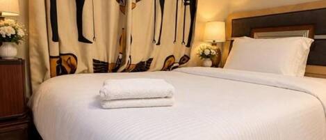 Cama bem arrumada com lençóis brancos, um par de chinelos no chão e cortina com padrão de pássaros.
