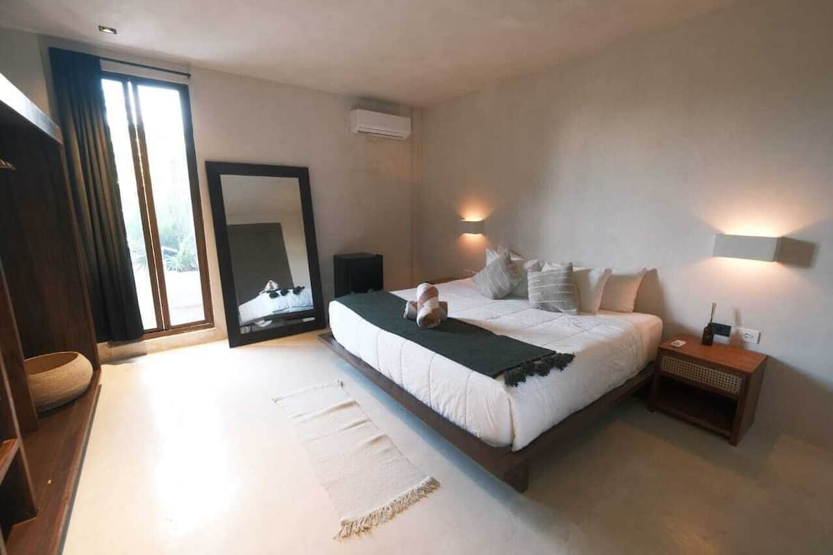 Quarto com uma cama grande, luminárias laterais, um espelho e uma janela com cortinas.